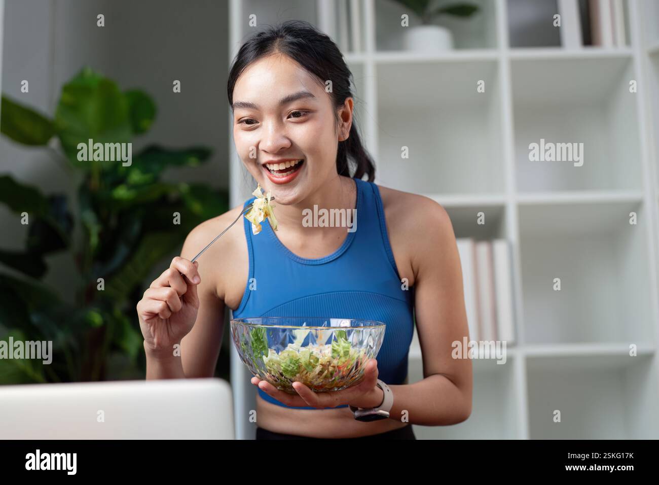 Excited woman laughing while eating salad and using a laptop, embracing ...