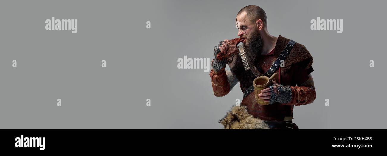 Man dressed as Viking warrior biting raw meat, holding wooden mug ...