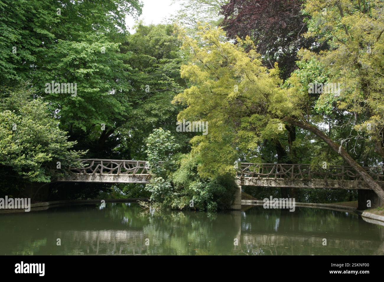Concrete bridge with intricate woven railings spans a tranquil pond ...