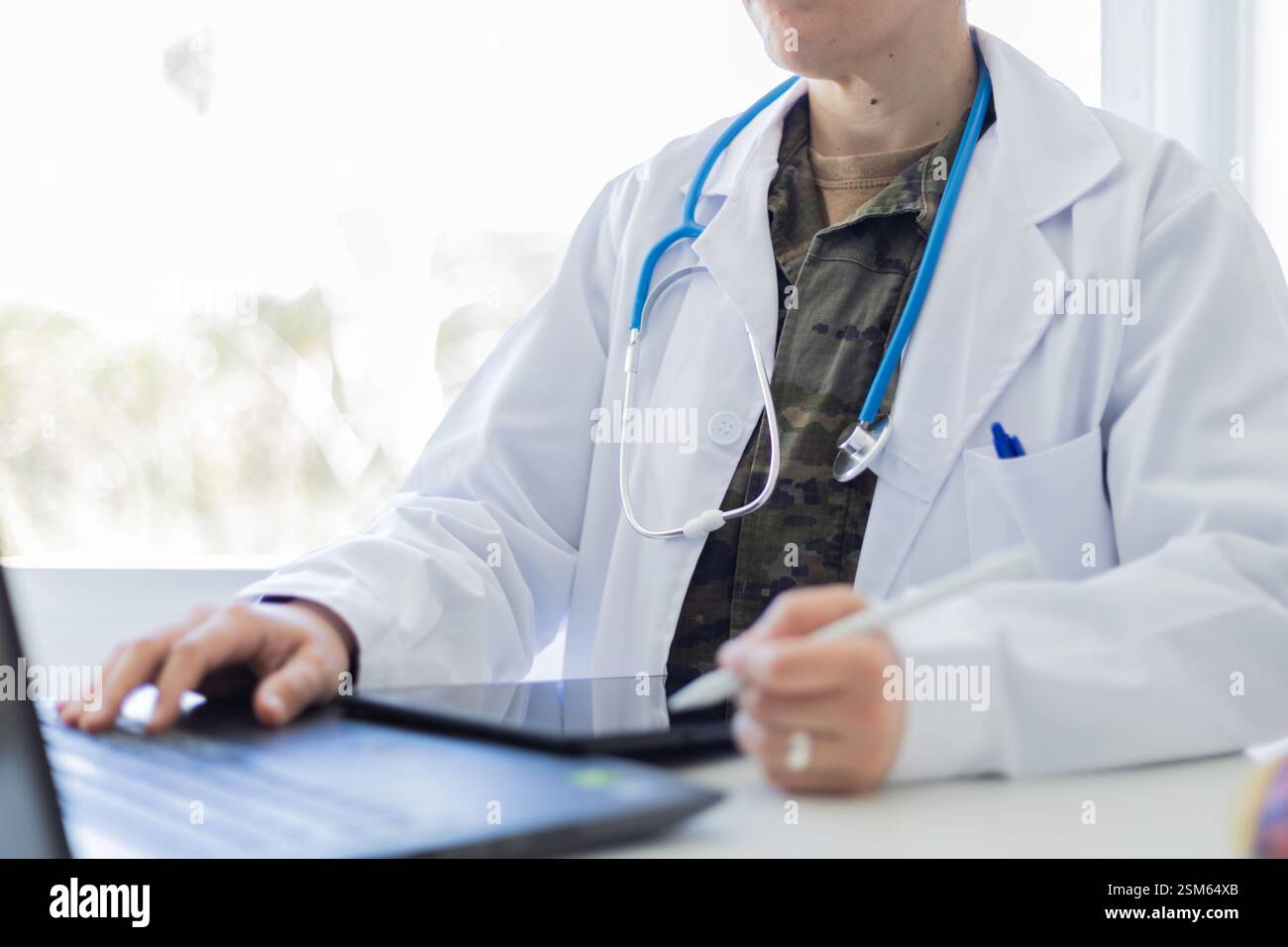 A military doctor wearing a white coat over a camouflage uniform works on a laptop and tablet in ...