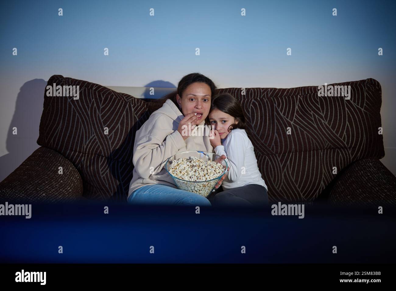 A mother and daughter sit on a cozy sofa, sharing popcorn while ...