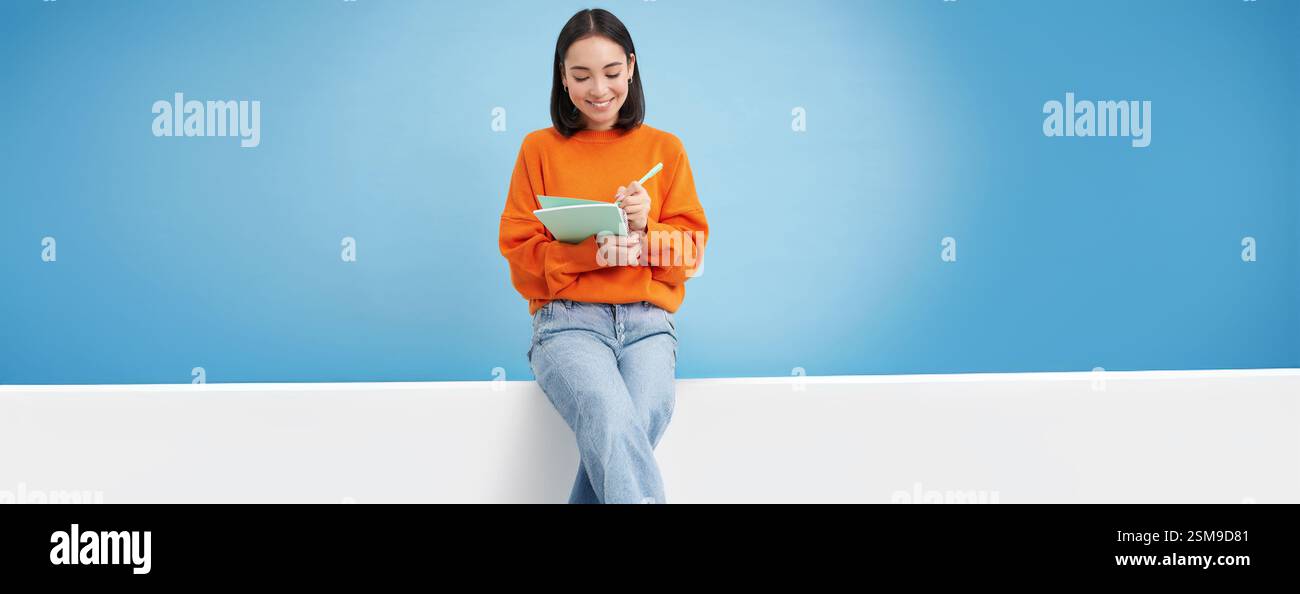 Smiling cute girl student, making notes, writing down homework ...