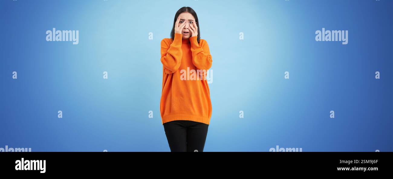Scared Asian woman shuts her eyes and peeks through hands at smth scary ...