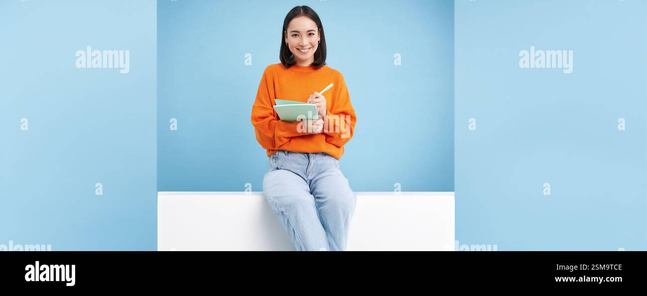 Smiling cute girl student, making notes, writing down homework ...