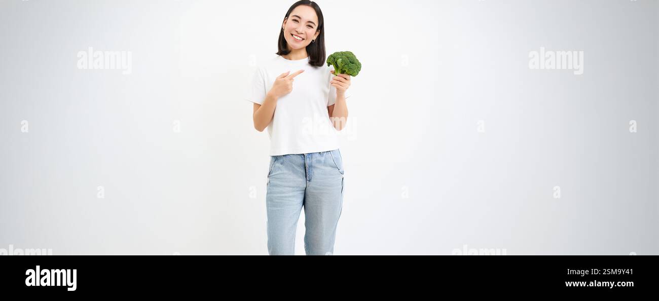 Portrait of cute asian woman, pointing at broccoli, showing vegetable ...