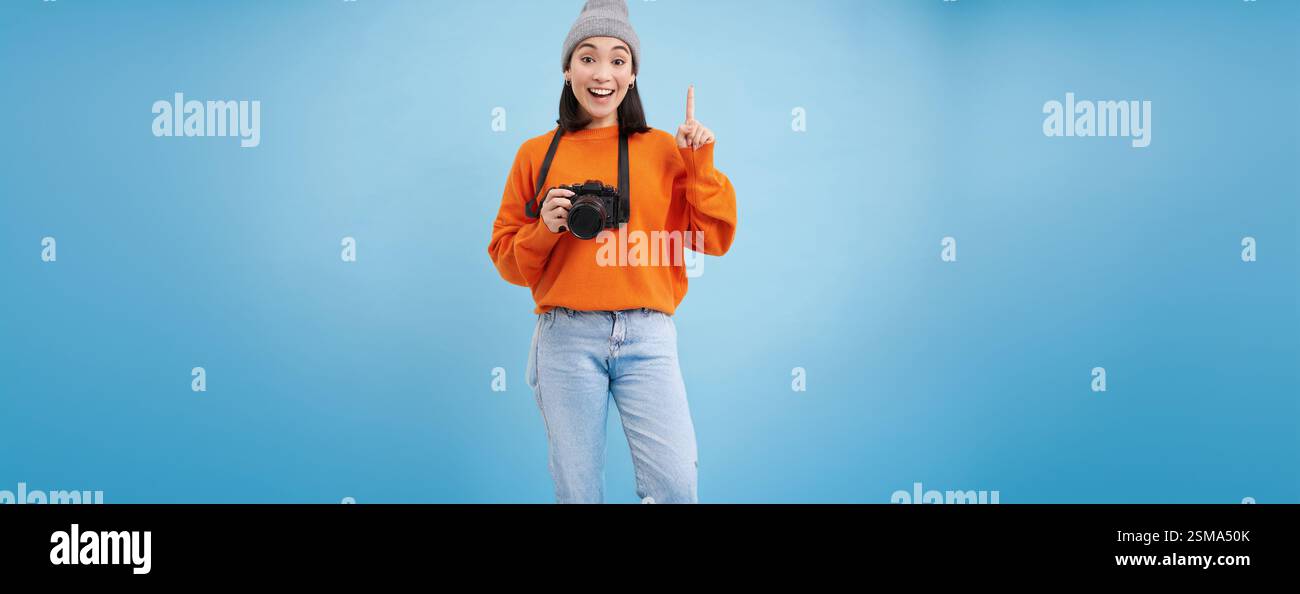 Surprised asian girl in hat, holds digital camera, looks amazed and ...