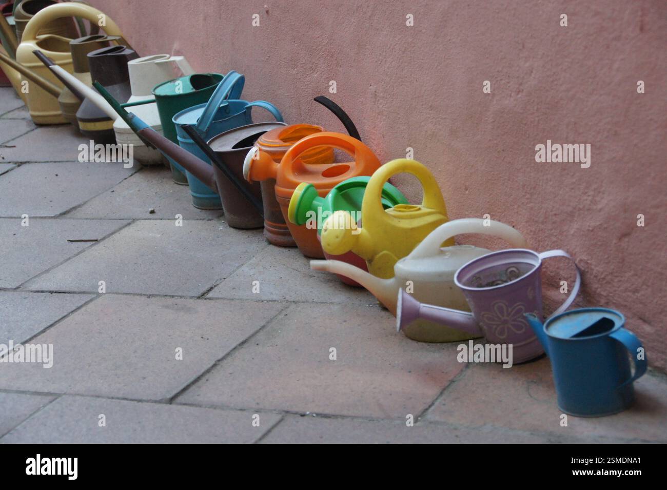 The photo shows a line of colorful watering cans lined up on a stone ...