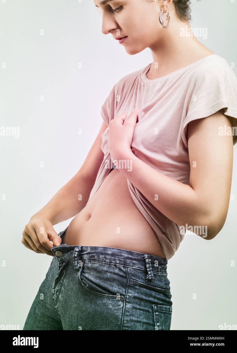 Young slim woman lifting her shirt and pulling up her jeans, proudly revealing her weight loss ...