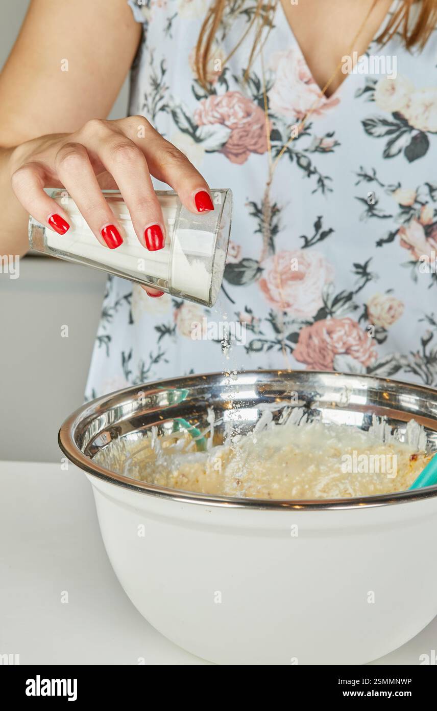 A woman pours flour into a metal mixing bowl, adding it to the batter ...