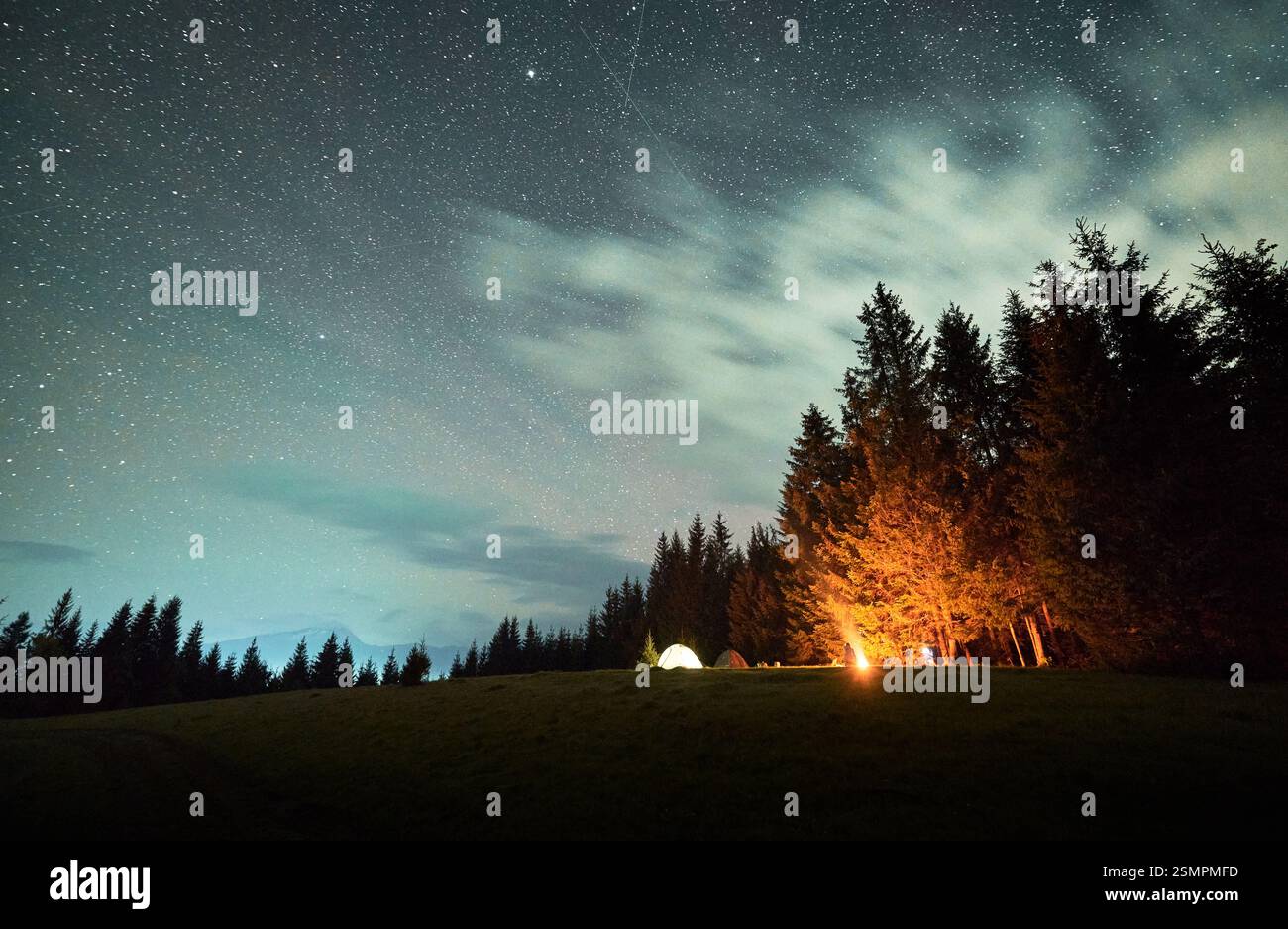 Campfire glows warmly beside several illuminated tents on grassy ...