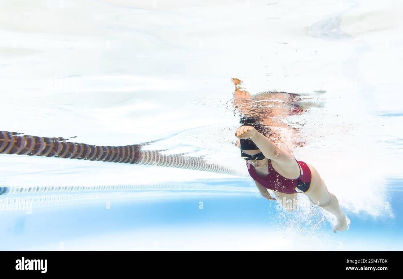 Female underwater swimmer in red swimsuit gliding through clear pool water, creating bubbles and ...