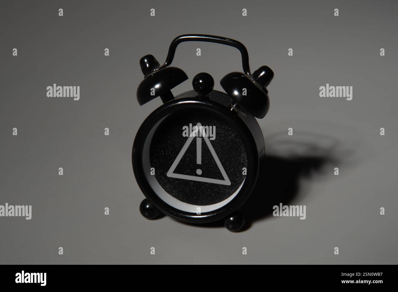 A black alarm clock with a white exclamation mark on a dark background ...