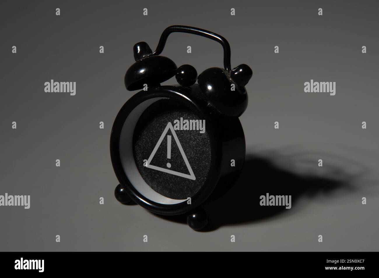 A black alarm clock with a white exclamation mark on a dark background ...