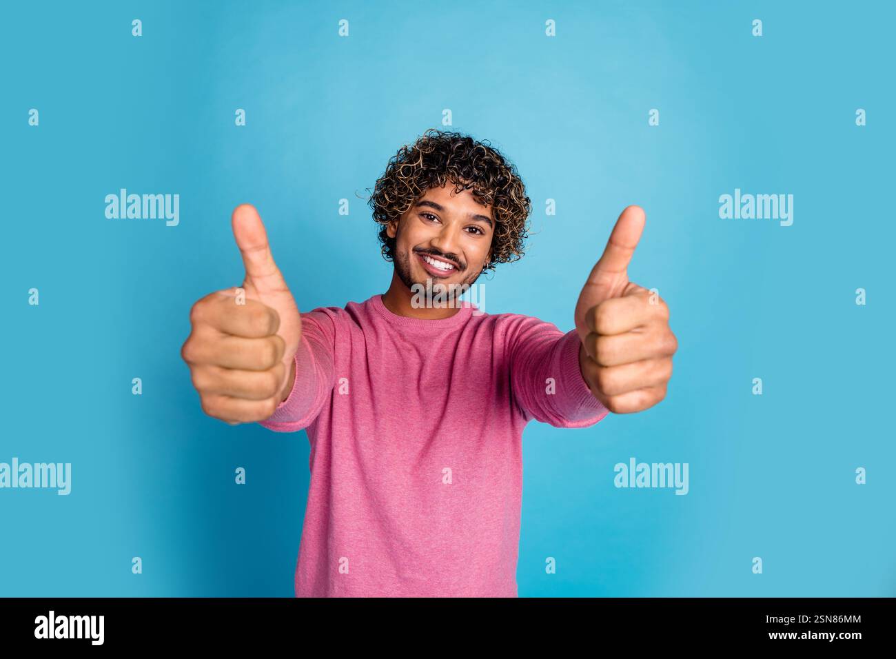 Smiling young man shows thumbs up gesture on blue background wearing ...
