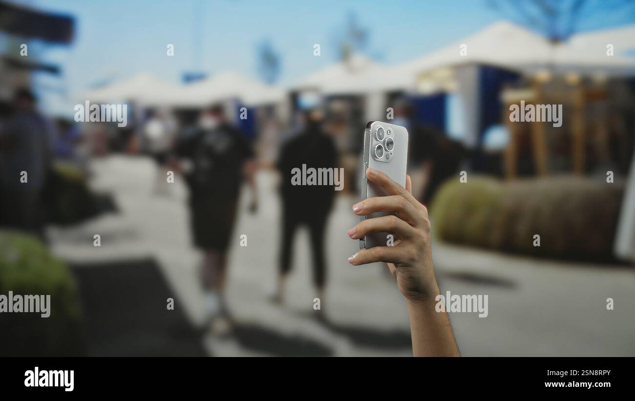 Hand holding smartphone outdoors at a bustling market with blurred ...