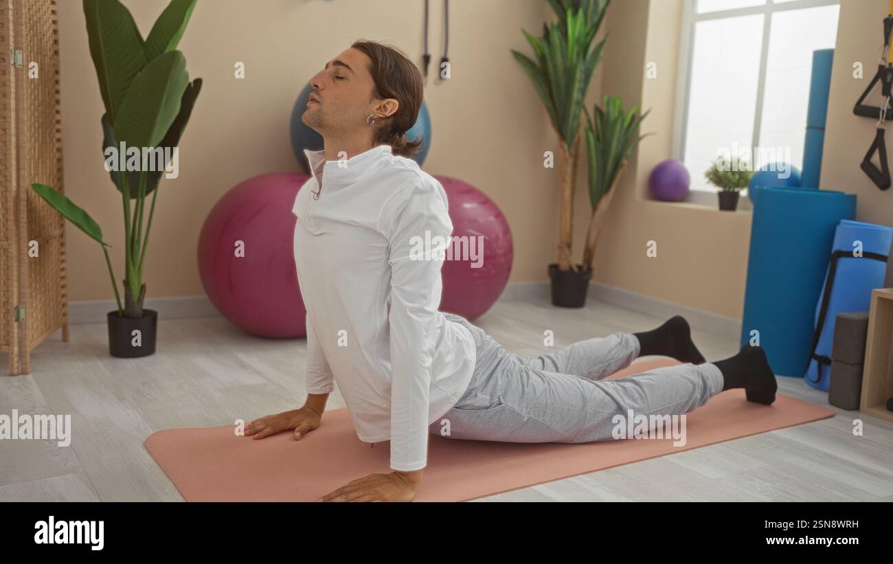 Young hispanic man with long hair practicing yoga in an indoor gym with ...