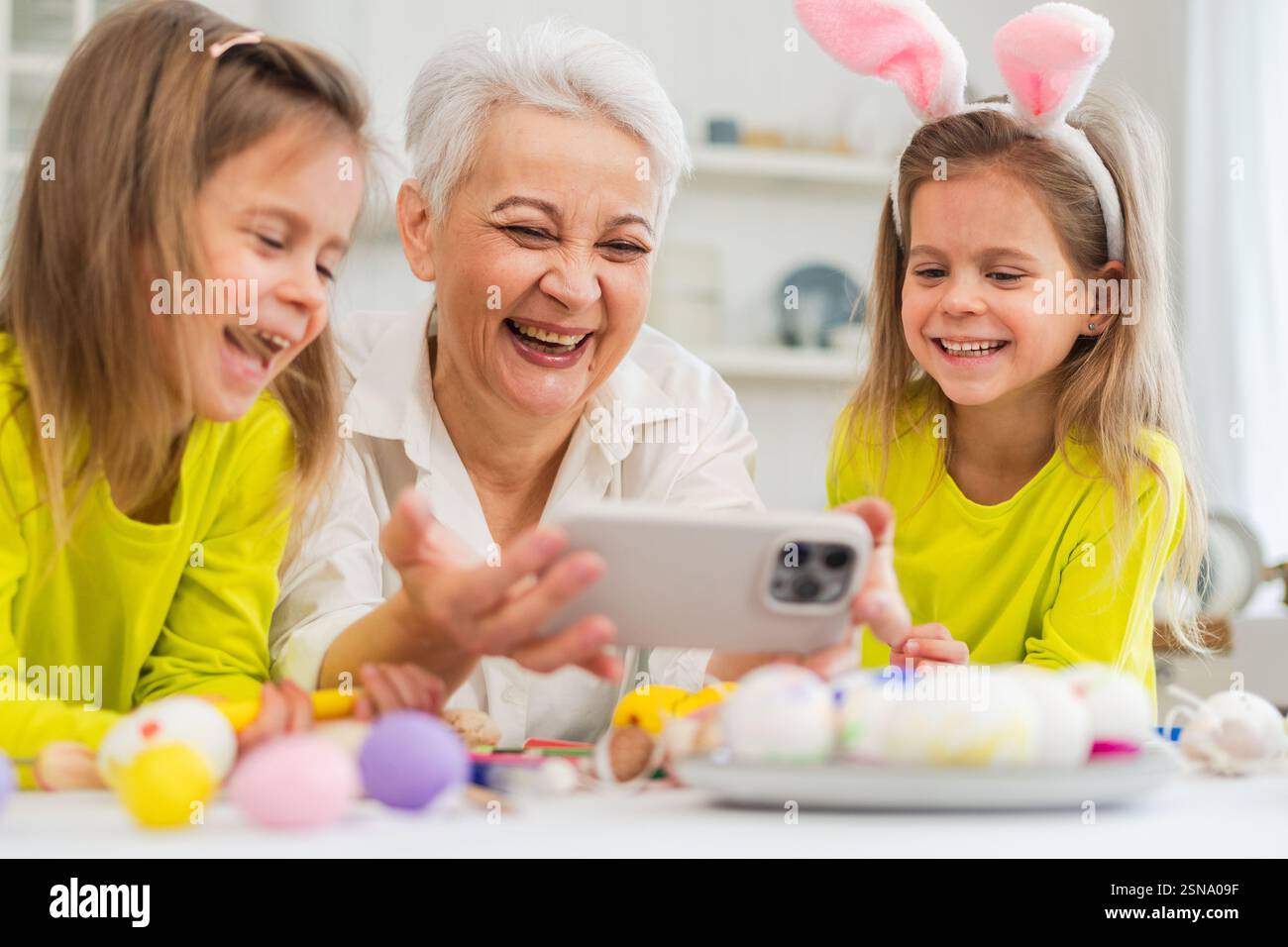 Happy Easter. Two girls sisters twins grandmother painting eggs watching video on phone enjoying ...