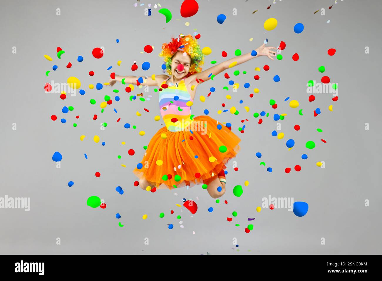 Happy girl dressed like clown jumping under falling confetti on grey ...