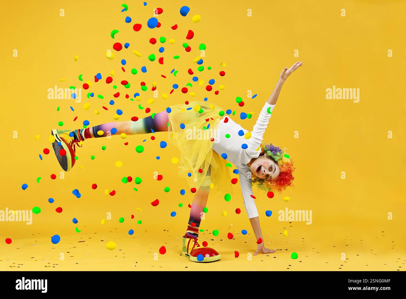 Happy girl dressed like clown under falling confetti on orange ...