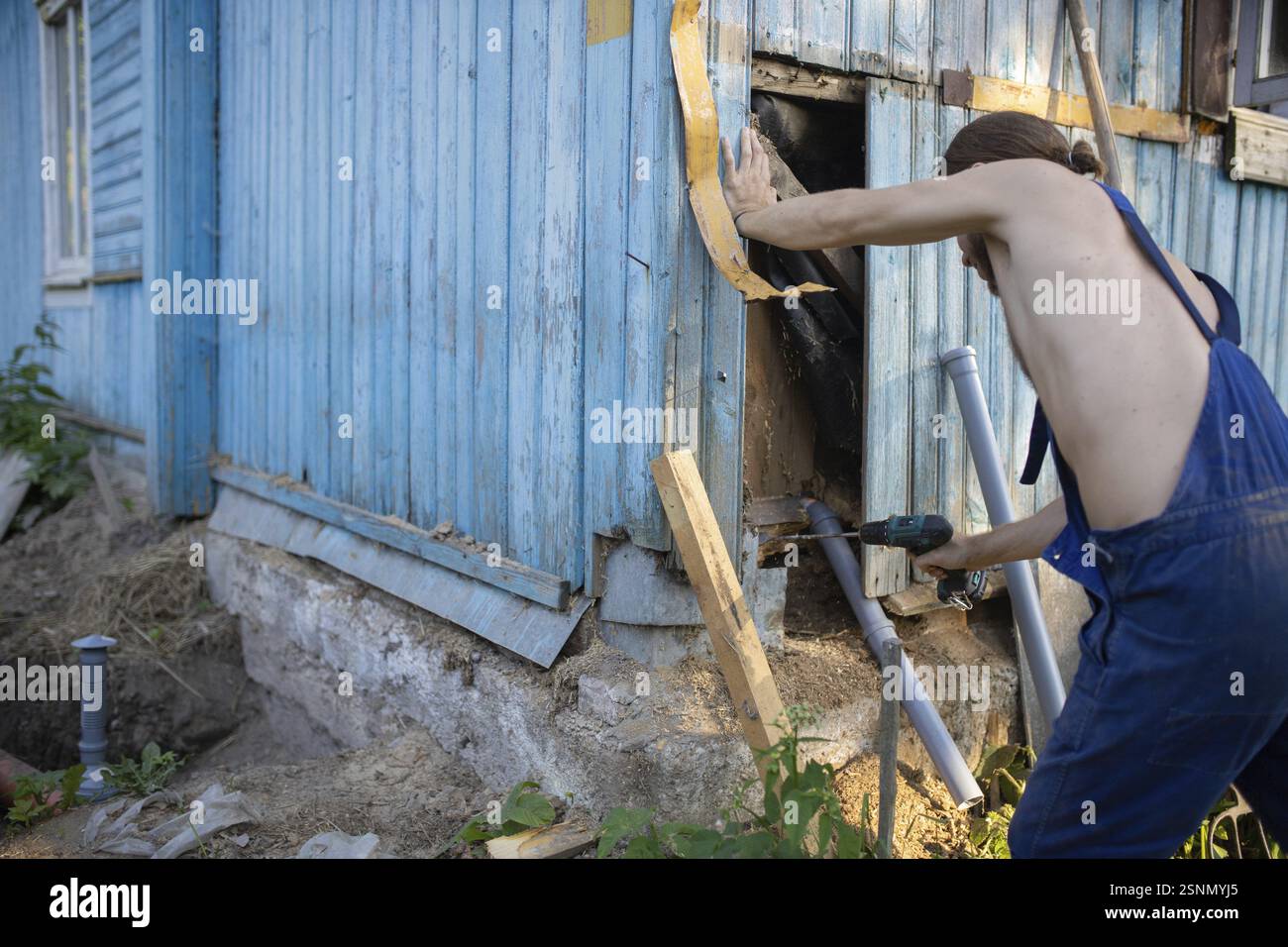 A man repairing an exterior wall of a wooden house in blue overalls ...
