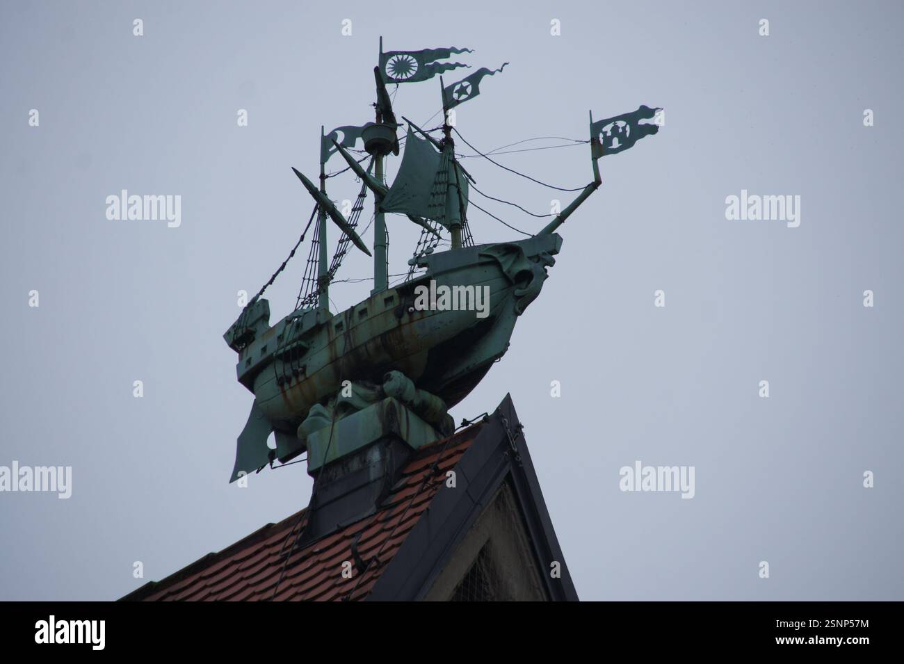 A weathered green ship weathervane with flags adorns a red tiled roof ...