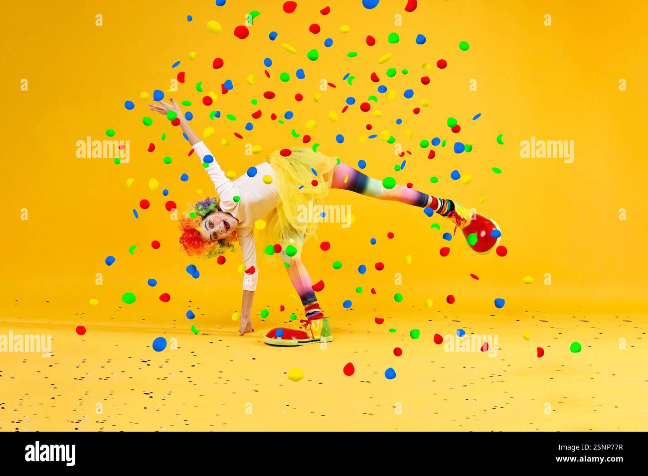 Happy girl dressed like clown under falling confetti on orange ...