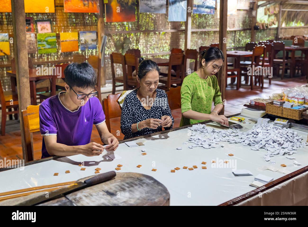 Three individuals focus on preparing traditional packs of coconut candy ...