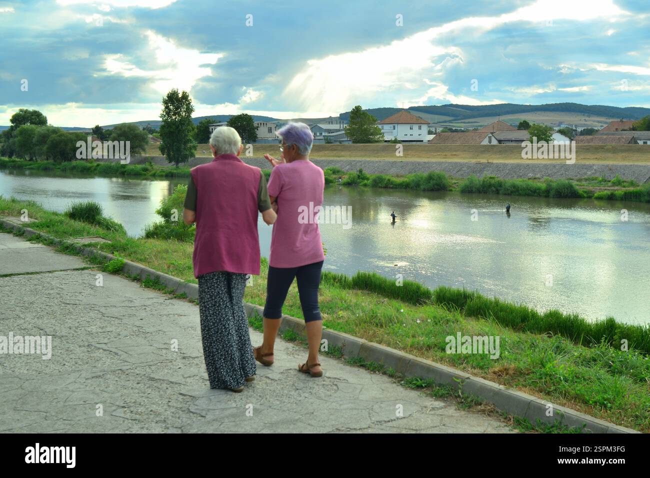 Two mature ladies walking and talking by the river with fishermen in ...