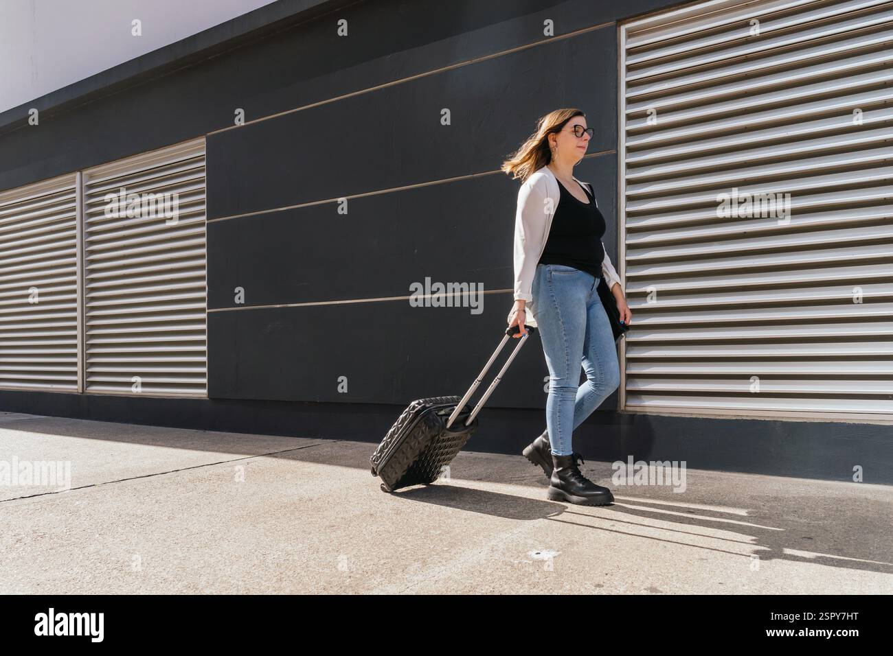 Businesswoman pulling trolley suitcase while walking on a sidewalk in ...