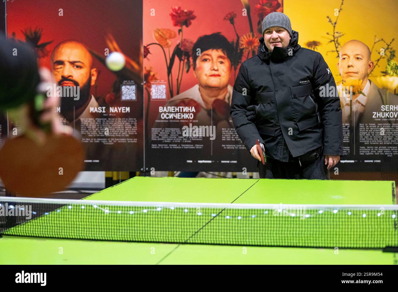 Visitors play ping-pong at a craft art fair as a poster of superhumans ...