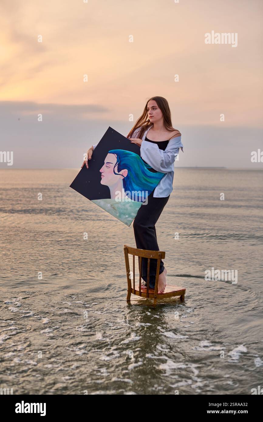 Young female painter stands on chair in serene water, holding vibrant ...