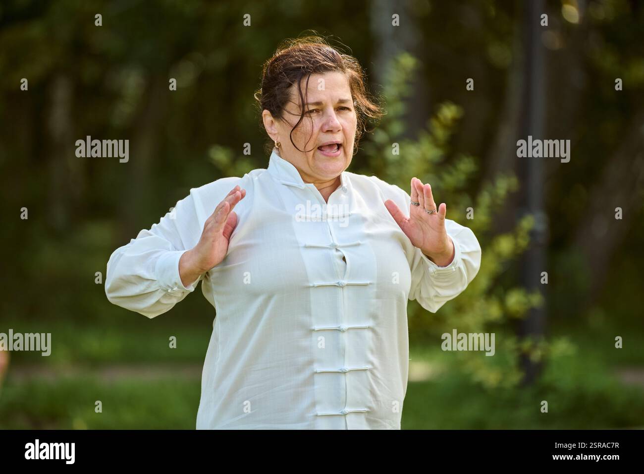 Mature woman with brown hair practices qigong outdoors, surrounded by nature. Wearing ...