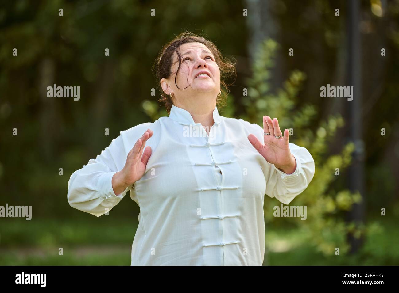 Mature woman with brown hair practices qigong outdoors, surrounded by nature. Wearing ...