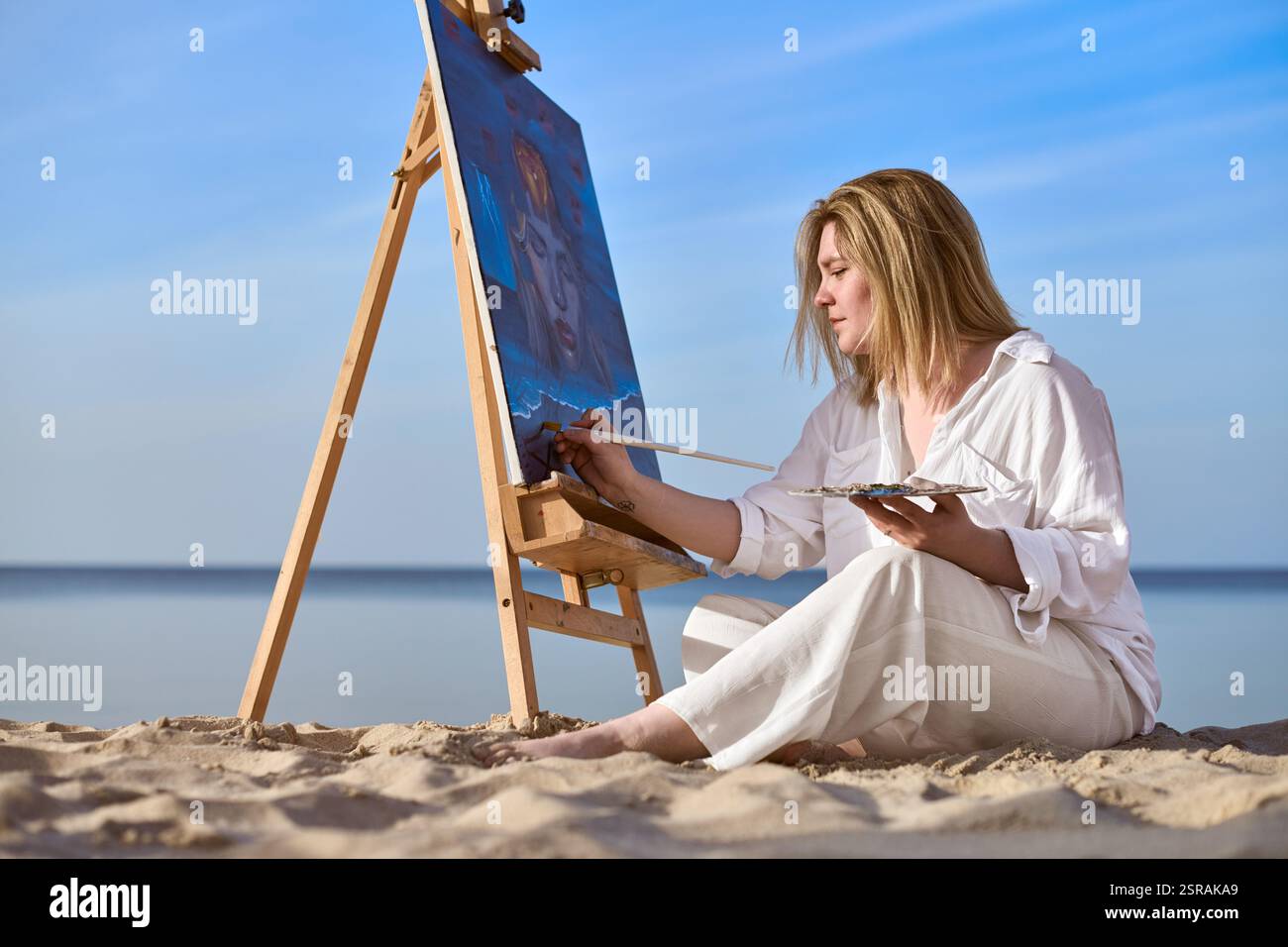 Young woman artist sits barefoot on tranquil beach, painting seascape ...