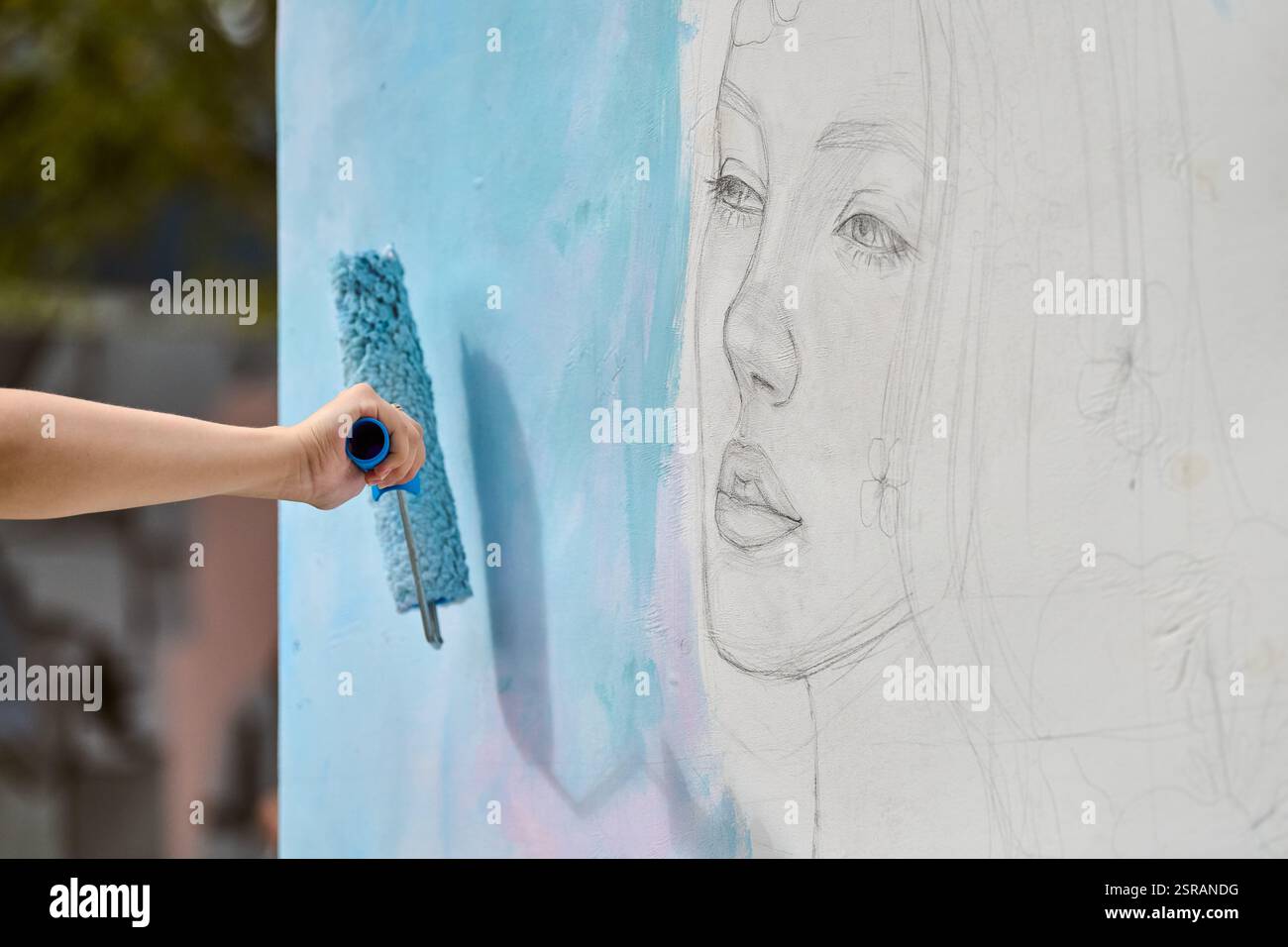 Hand applies soft blue paint over pencil sketch of young female face on white wall. Natural ...