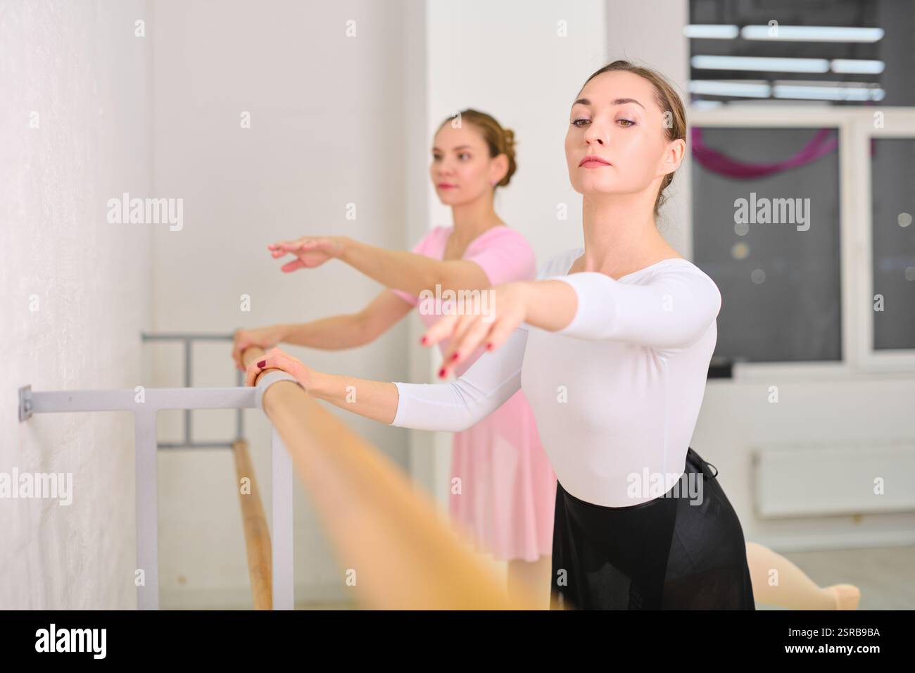 Two young women with light brown hair practice ballet at barre in ...