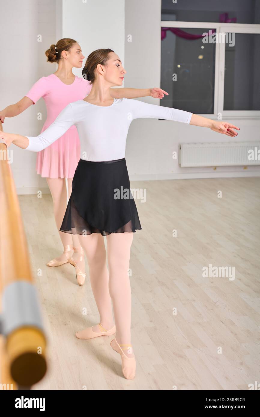 Two young female ballerinas with brown hair rehearsing at ballet barre ...