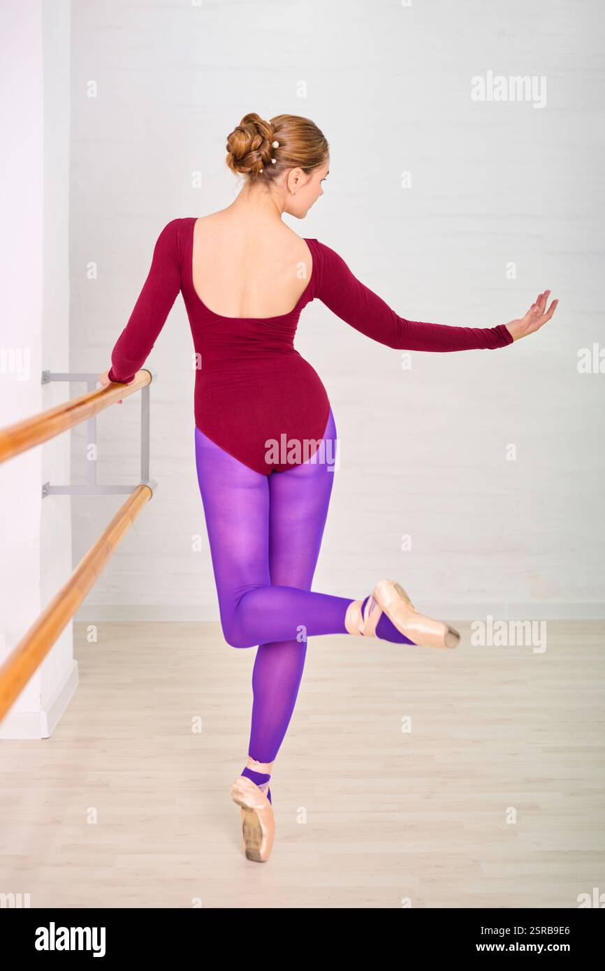 Young female ballerina with auburn hair in bun practices at barre ...
