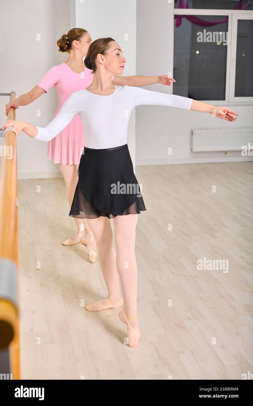 Two young female ballerinas with brown hair rehearsing at ballet barre ...