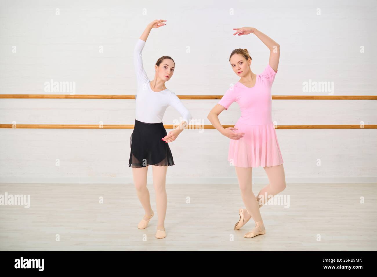 Two young adult female ballerinas with light brown hair practice ballet positions at barre in ...
