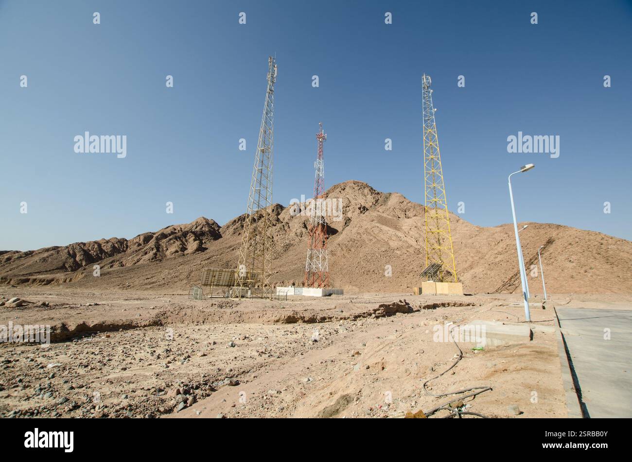 Tall communication towers stand amidst barren, rocky terrain under clear blue sky. Sunlight ...