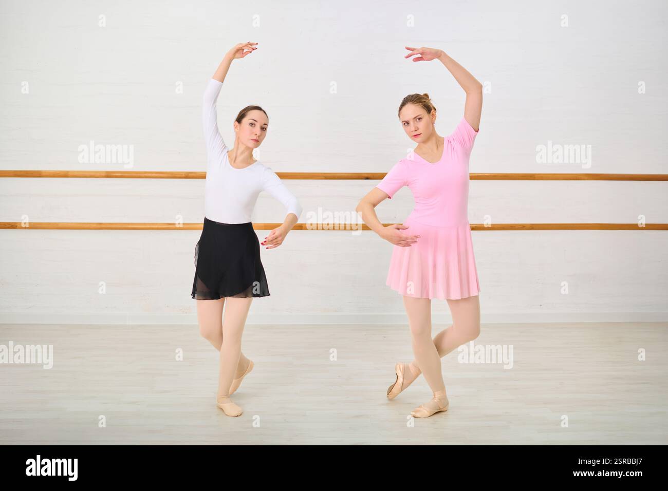 Two young adult female ballerinas with light brown hair practice ballet positions at barre in ...