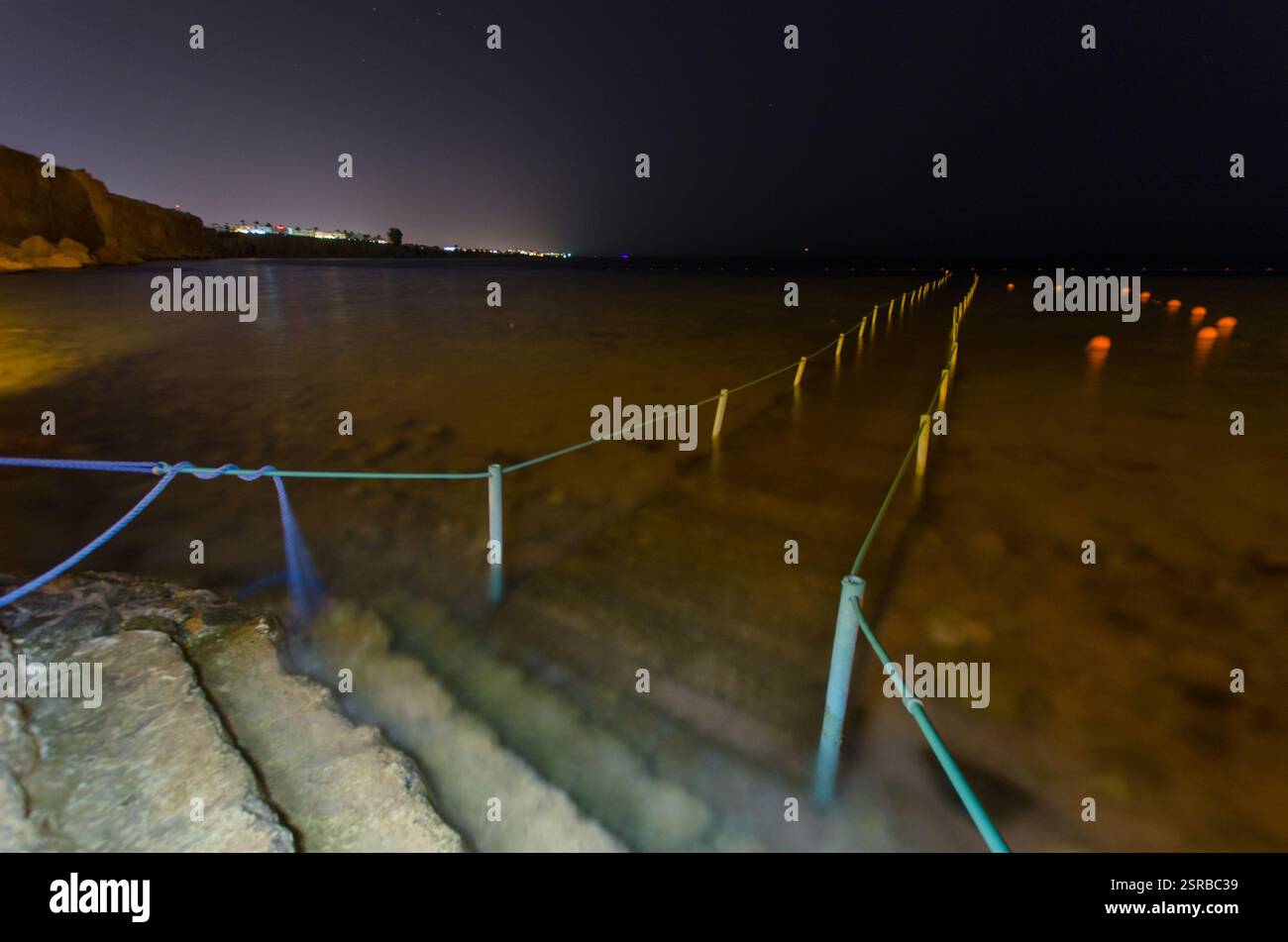Dimly lit pathway leads into calm night waters at resort beach. Soft ...