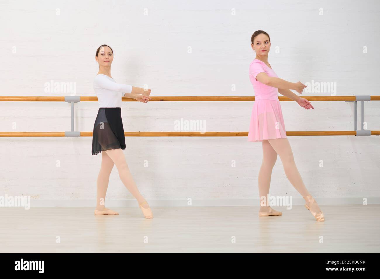 Two young female ballerinas with dark hair practice ballet at barre ...