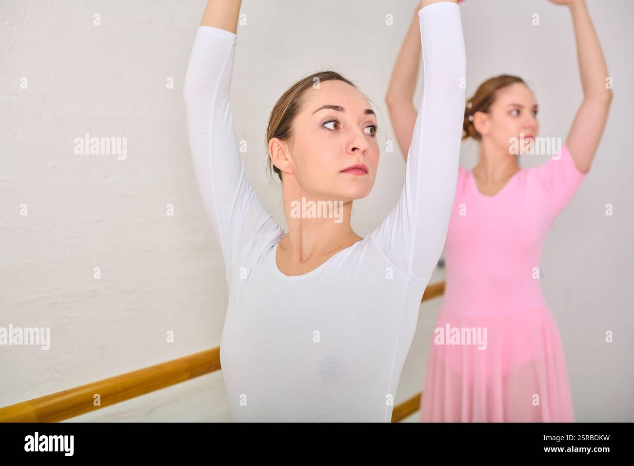 Two young female ballerinas practice at ballet barre in bright studio. One in white leotard ...