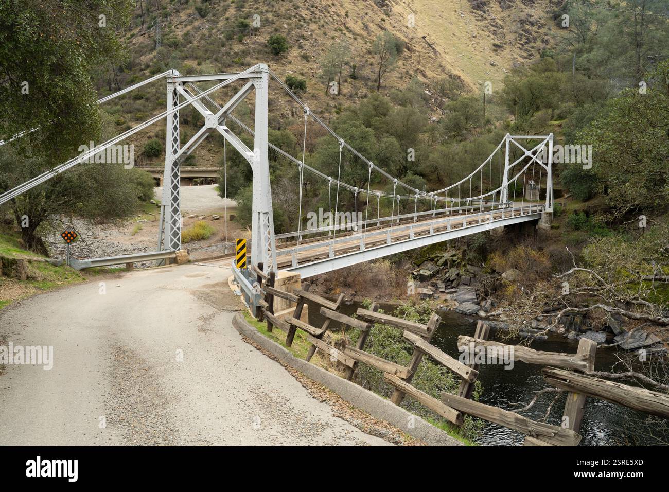 Old suspension bridge spans a river. Road curves beneath, worn wooden ...