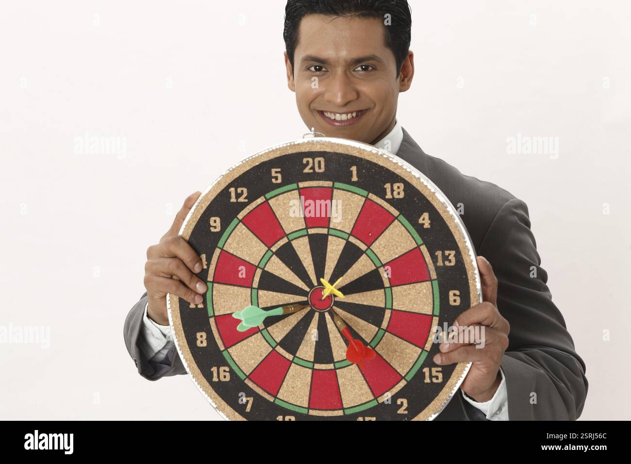 Businessman holding dart board in front of his chest showing three darts on centre in both hands ...
