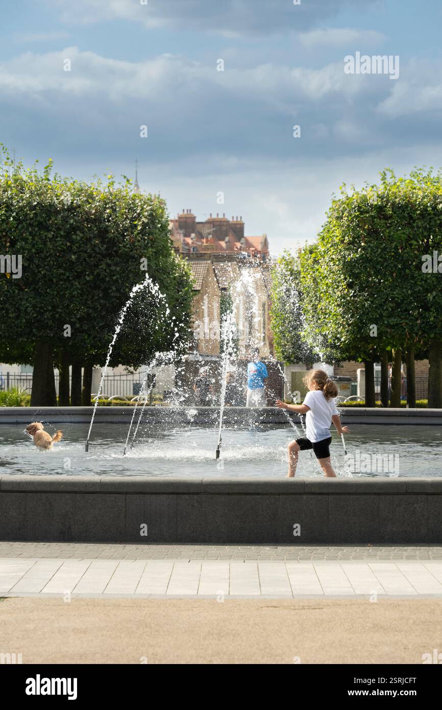 Child plays in the fountain at the Imperial Park, London. A dog also ...