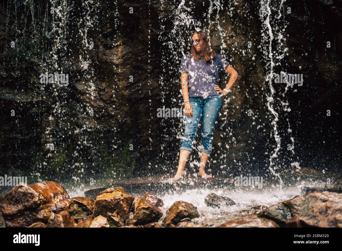 A woman in casual clothes stands barefoot behind a cascading waterfall, surrounded by rocky ...