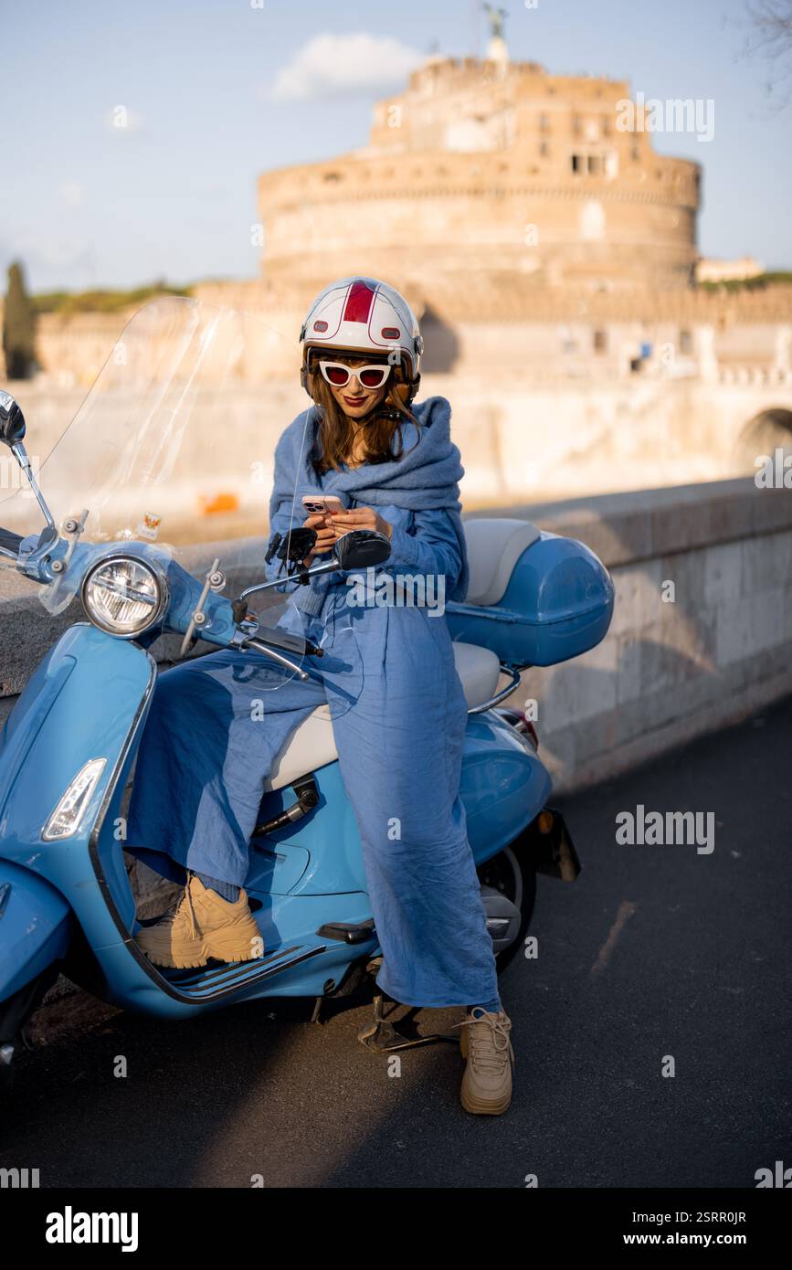 Stylish woman in a blue outfit and helmet sits on a scooter, checking ...
